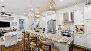 Uma cozinha moderna com armários brancos, bancada de granito e geladeira de aço inoxidável. Três luminárias pendentes de vime iluminam a ilha.