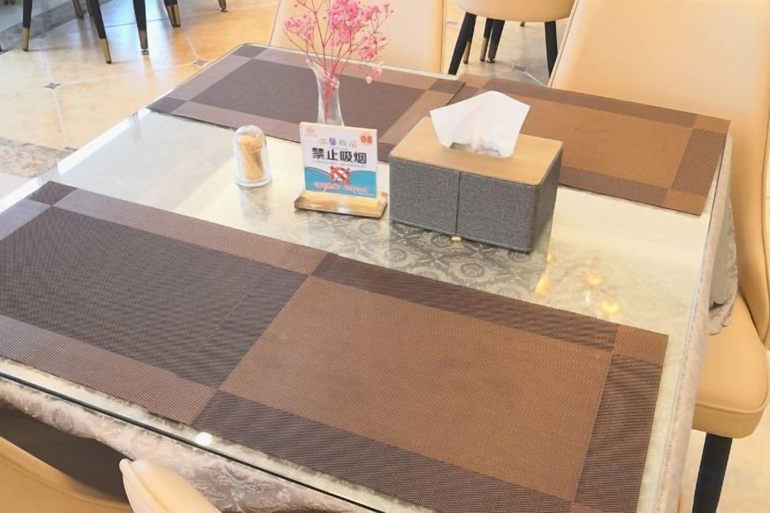 Quarto de hotel com mesa de vidro, caixa de lenços e pequeno vaso com flores cor-de-rosa.