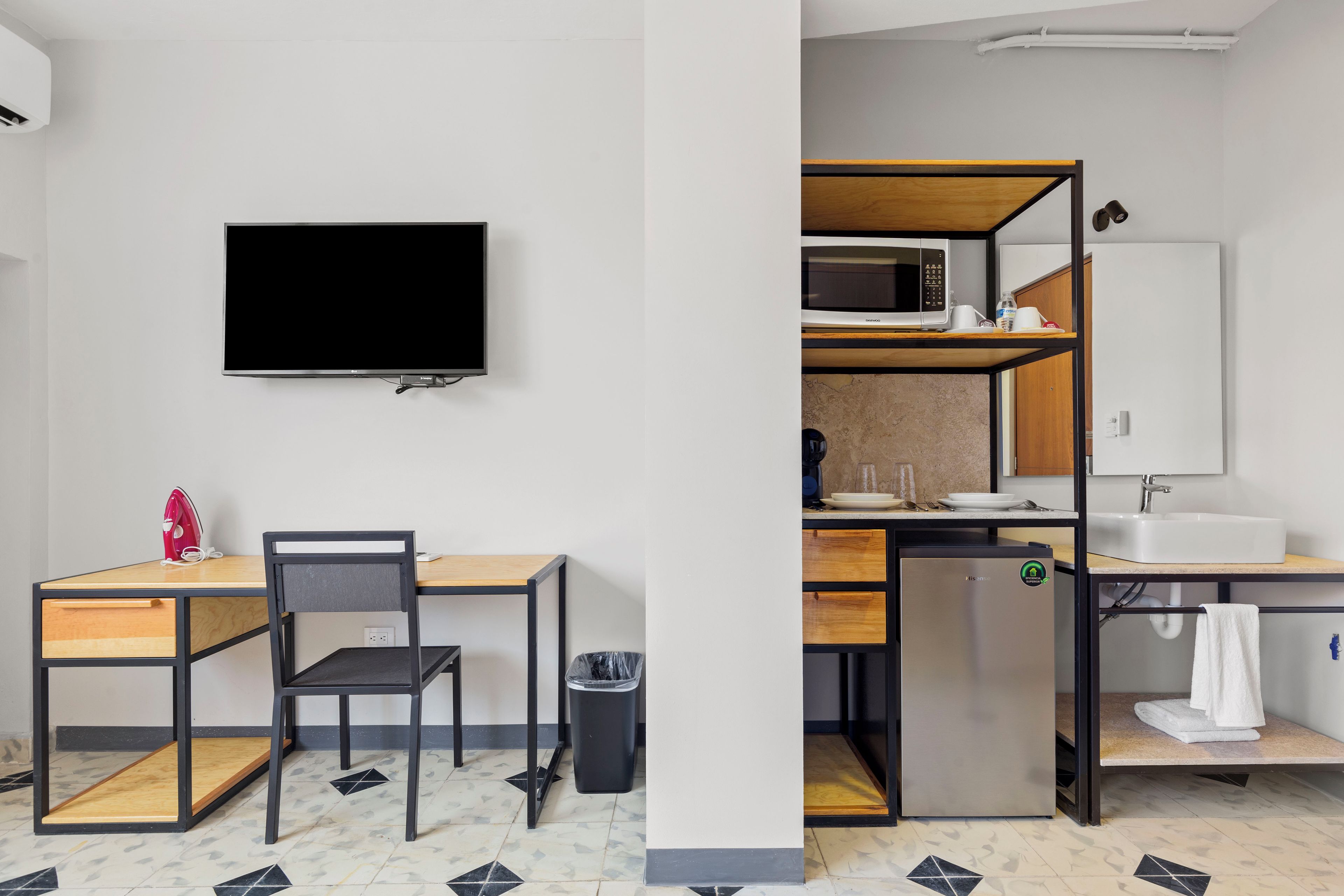 Quarto compacto com cama, escrivaninha e cadeira, televisão, micro-ondas, pia e mini-geladeira.