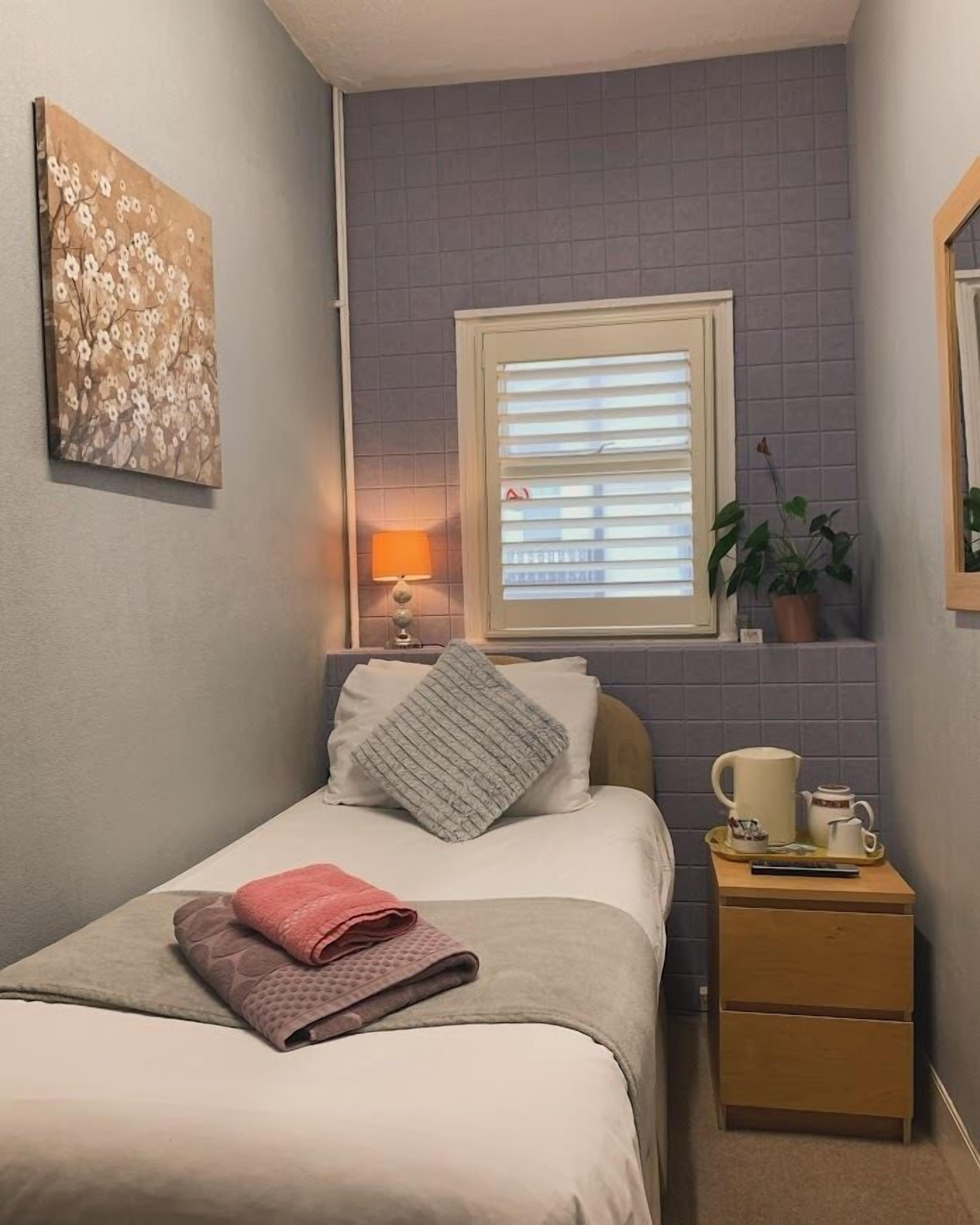 Quarto bem organizado com cama, mesa de cabeceira, abajur e janela com persianas.