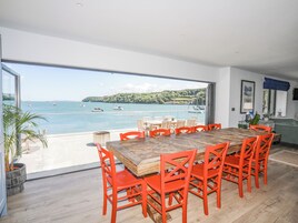 Uma área de refeições com uma mesa grande e cadeiras laranja, com vista para uma baía com barcos e uma paisagem montanhosa.