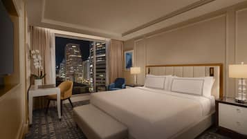 Quarto de hotel com uma cama grande, uma escrivaninha, uma cadeira, uma mesinha de cabeceira e vista para a cidade à noite.