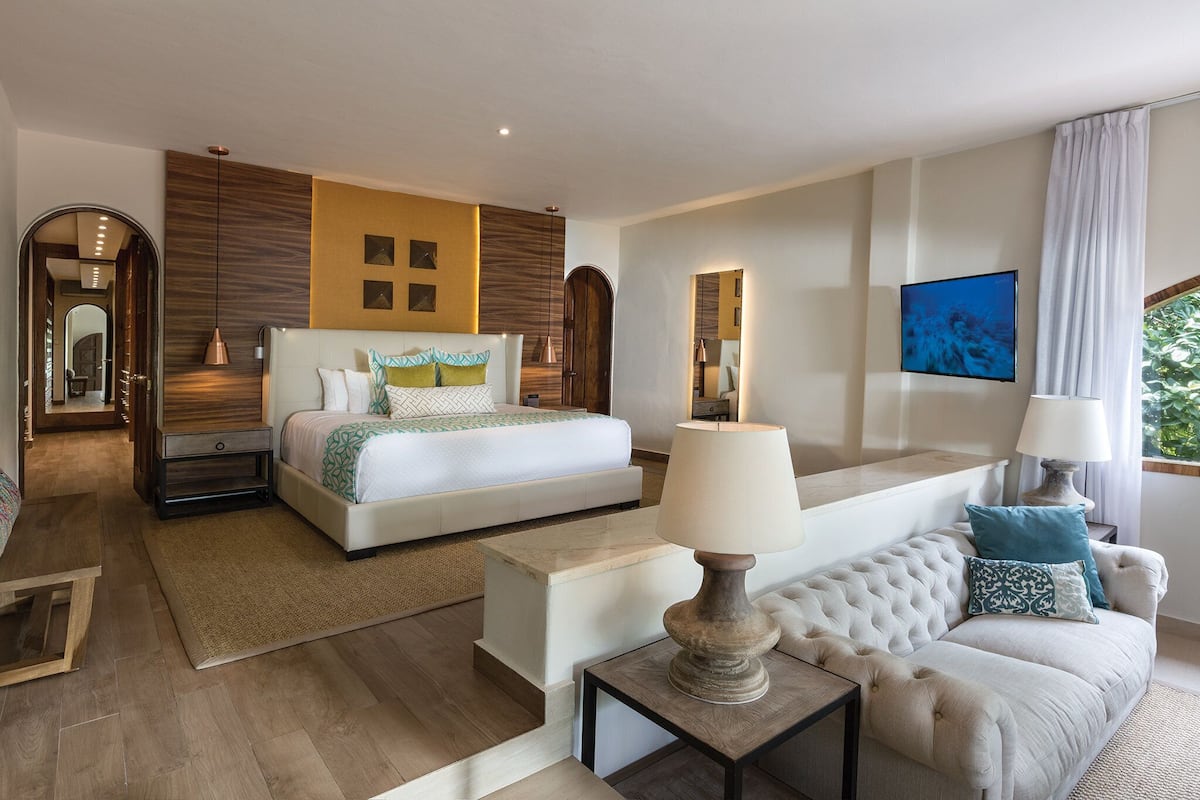 Quarto de hotel moderno com uma cama grande, um sofá, uma luminária e uma televisão.