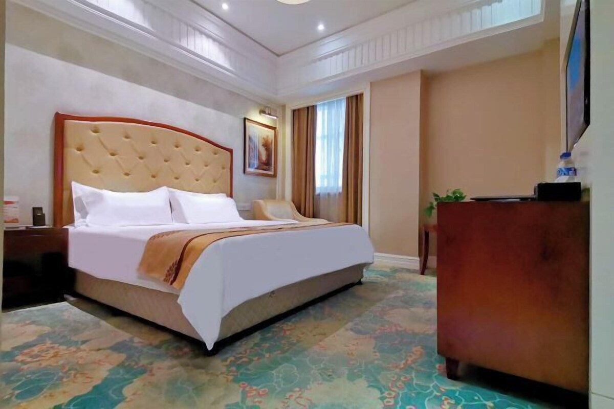 Quarto de hotel com uma cama grande, mesa de cabeceira de madeira, escrivaninha e televisão.