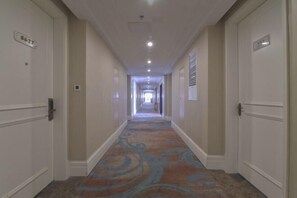 Corredor de hotel com piso coberto por carpete, portas brancas e uma placa de número em uma delas.