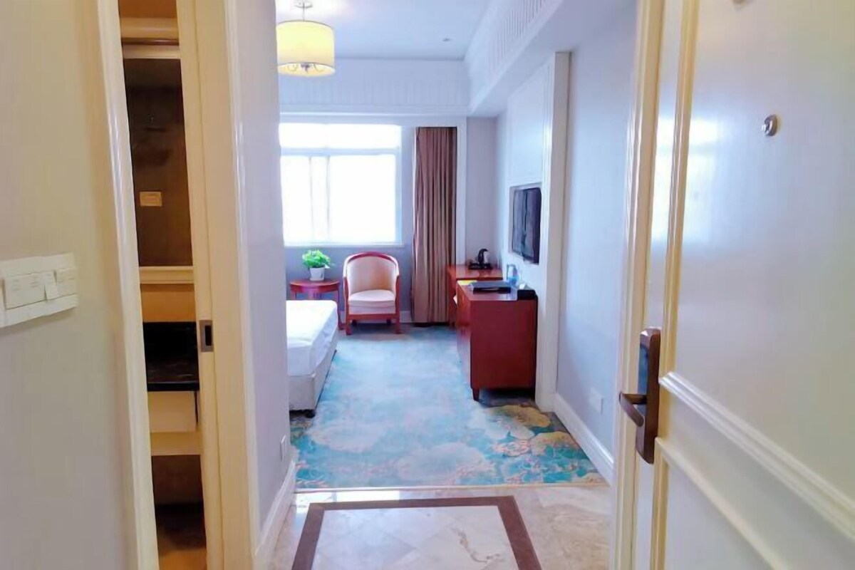 Quarto de hotel com cama, escrivaninha, cadeira, televisão e janela com cortinas.
