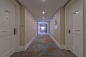 Corredor de hotel com piso coberto de carpete, portas em ambos os lados e luminária no teto.