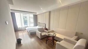 Quarto de hotel moderno com uma cama grande, dois sofás brancos, uma mesinha redonda e vista da cidade através de grandes janelas.