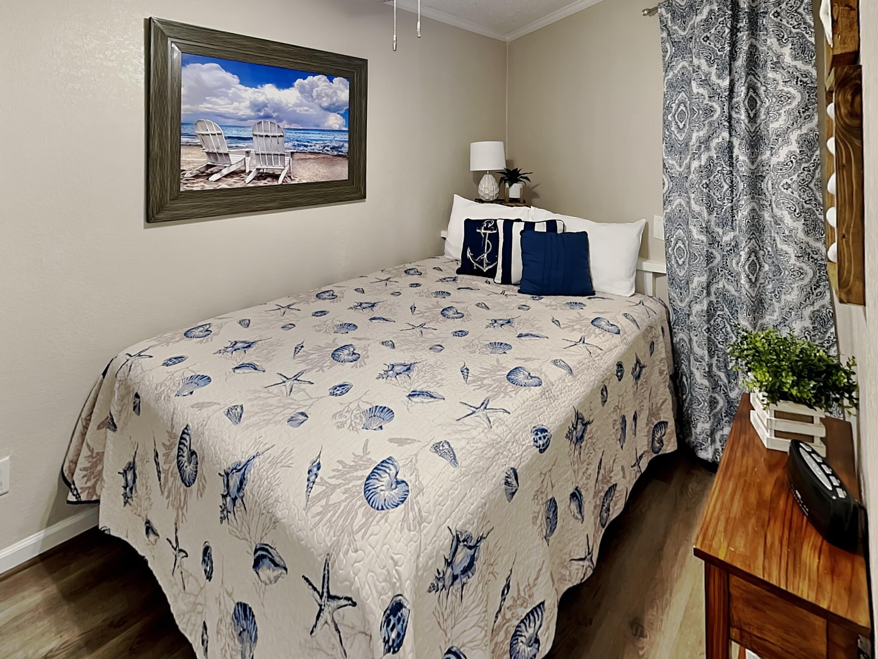 Quarto com cama, mesa de cabeceira, abajur, quadro na parede e janela com cortinas.