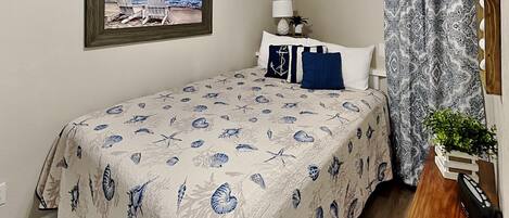 Quarto com cama, mesa de cabeceira, abajur, quadro na parede e janela com cortinas.