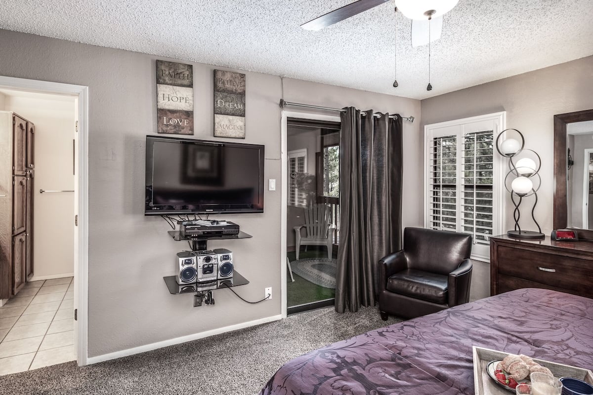 Quarto com cama, televisão na parede, poltrona de couro e janela com cortinas.