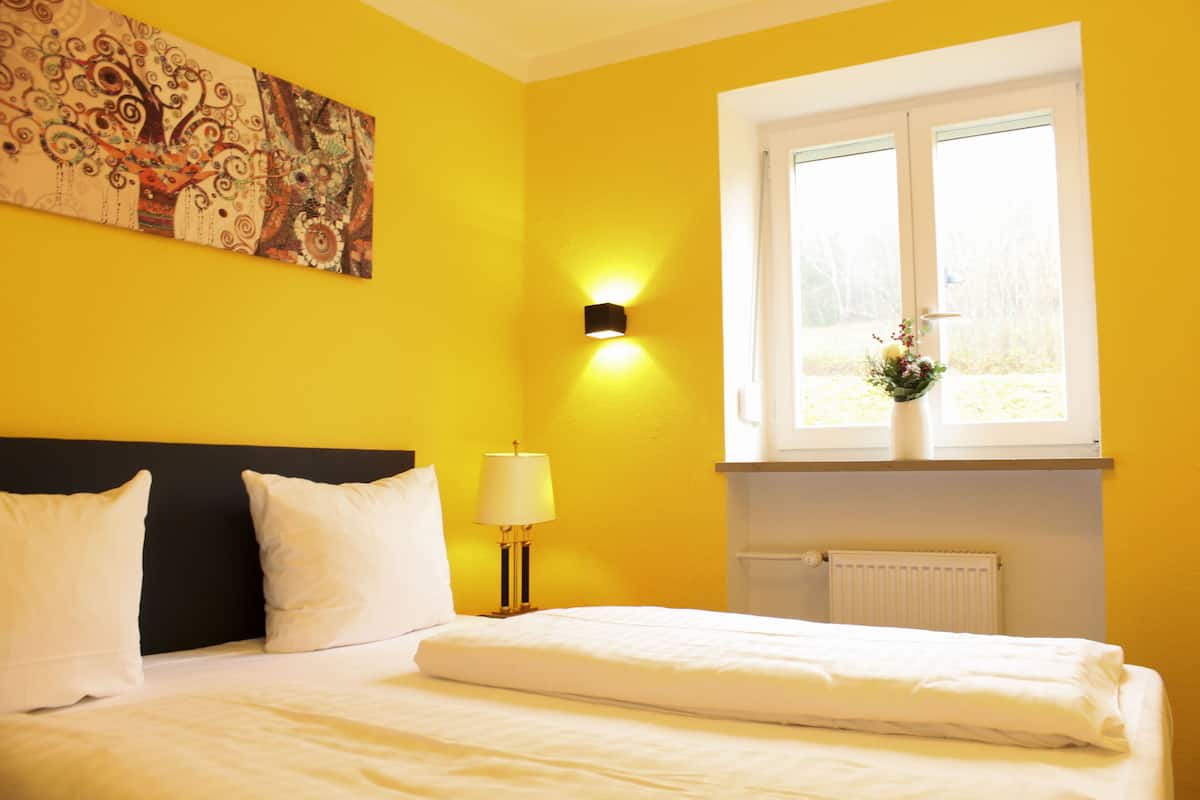Quarto de hotel com cama, roupa de cama branca, cabeceira preta, luminária na parede e janela com vista para árvores.