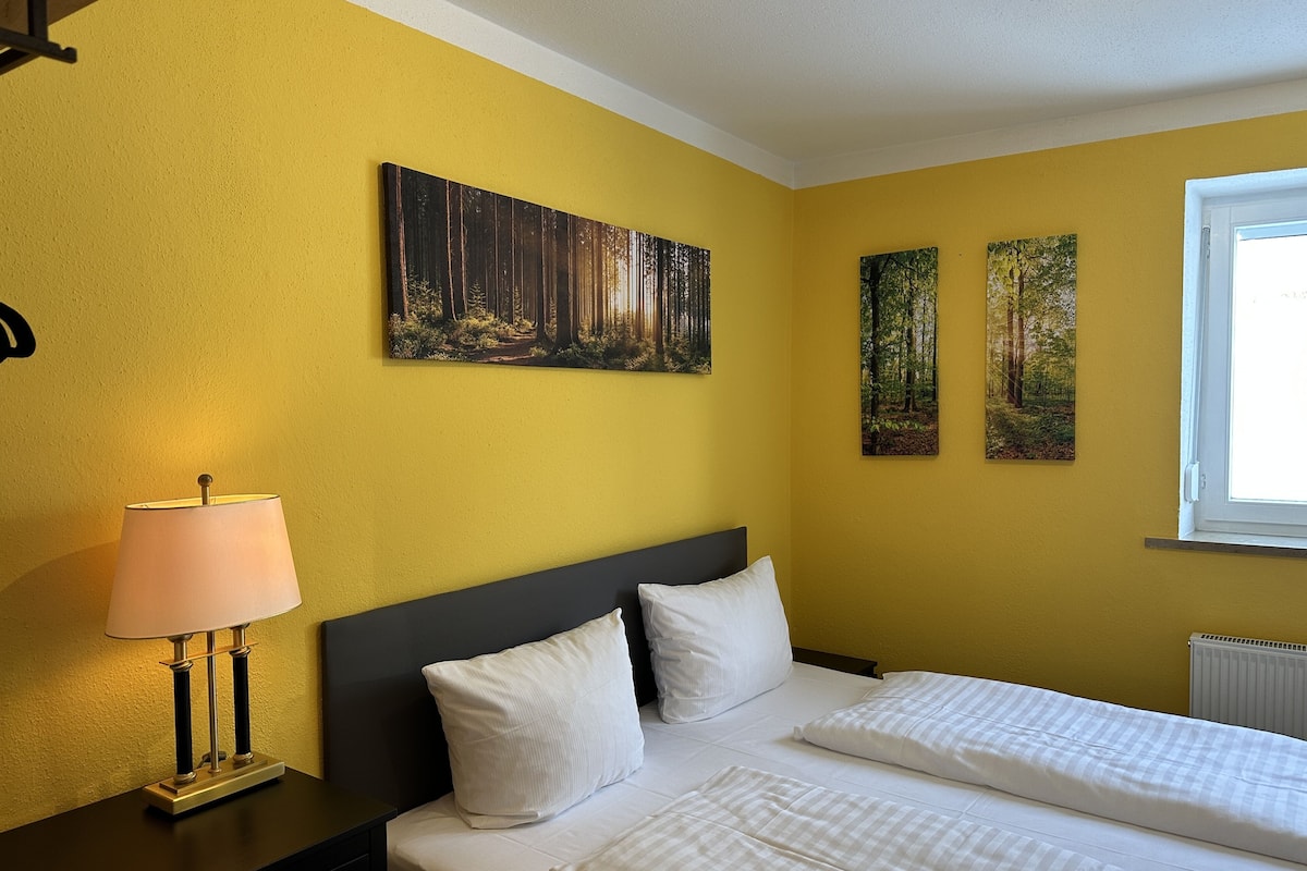 Quarto com cama, duas almofadas, mesa de cabeceira, abajur e duas pinturas penduradas na parede.