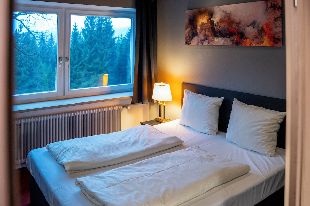 Quarto de hotel com uma cama, uma janela com vista para árvores, um quadro na parede e uma mesa de cabeceira com abajur.
