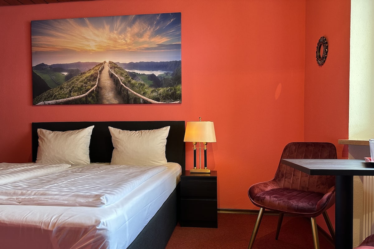 Quarto de hotel com uma cama, escrivaninha, cadeira e um quadro de uma paisagem na parede.