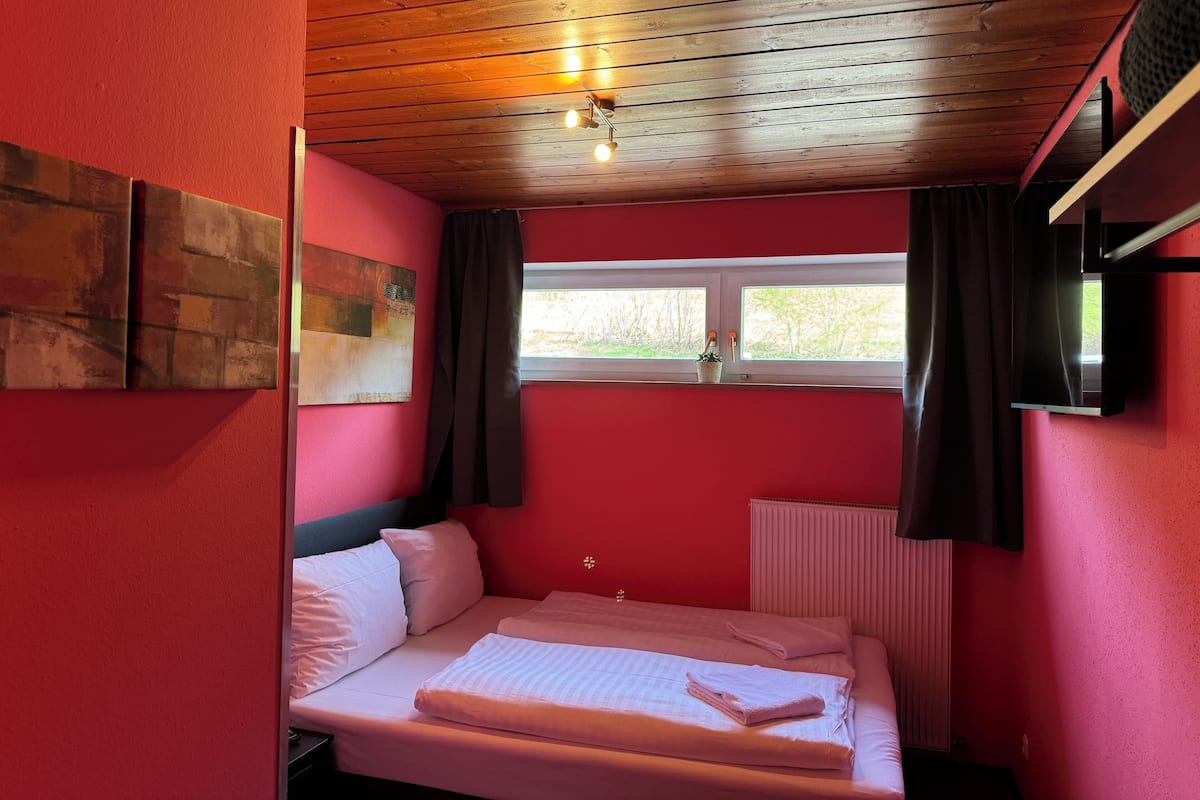 Quarto com teto de madeira, uma cama de solteiro com roupa de cama branca, janela com cortinas e um radiador.