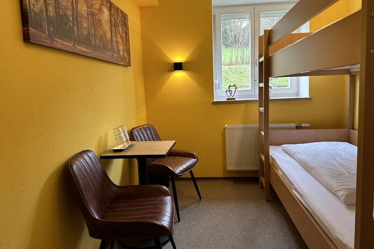 Quarto com cama de beliche, uma mesa com cadeiras, um quadro na parede e uma janela com vista para árvores.