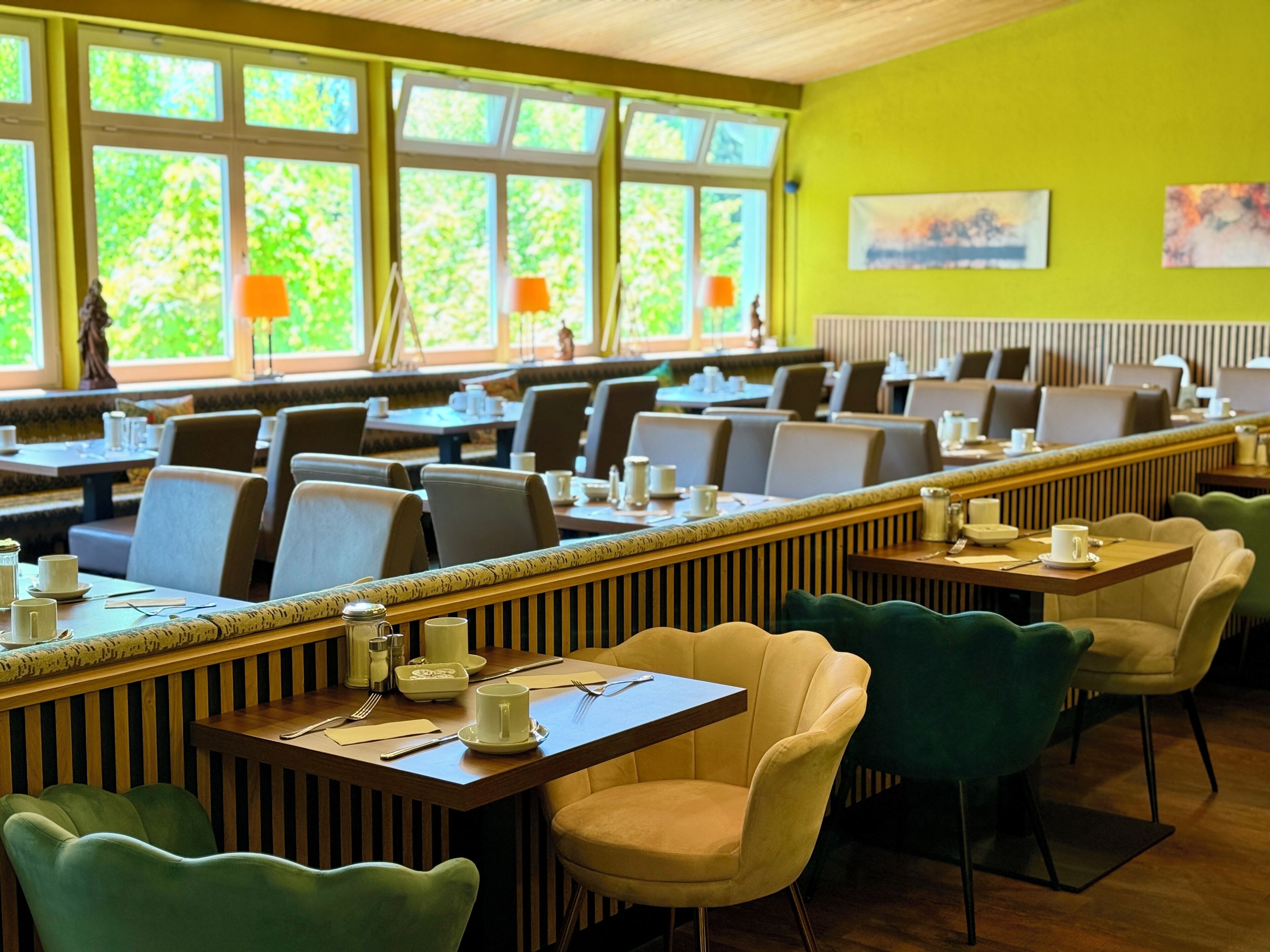 Um restaurante com várias mesas e cadeiras, grandes janelas com vista para o verde e uma parede amarela com um quadro.