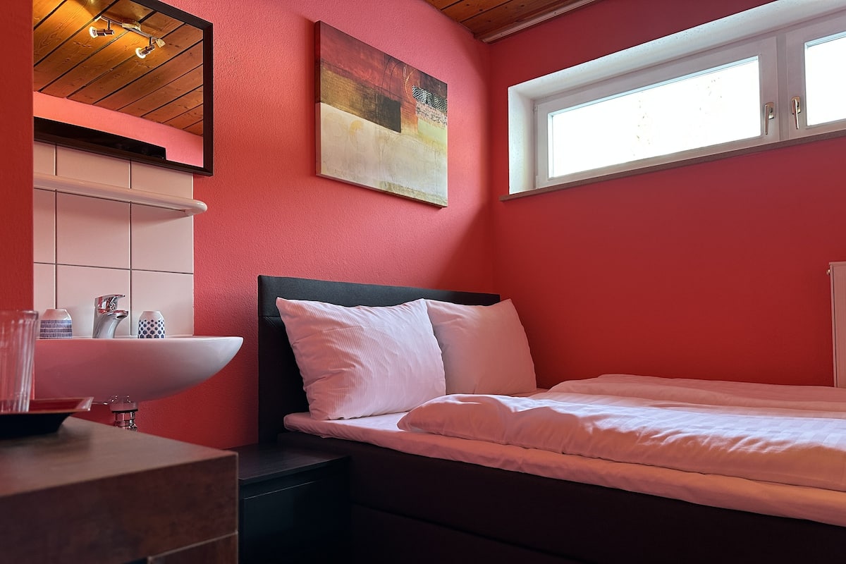 Quarto com uma cama, mesa de cabeceira com lavatório branco, um quadro na parede e uma janela.