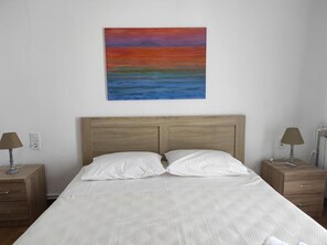 Quarto com cabeceira de madeira, roupa de cama branca e pintura de um pôr do sol sobre o oceano na parede.