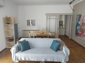 Uma sala de estar com sofá, mesa de jantar e cadeiras. Há uma estante de livros e uma pintura na parede.