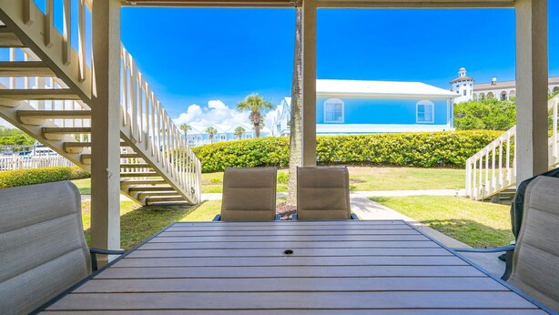 Uma área de estar coberta ao ar livre, com mesa e cadeiras, uma escadaria e vista para um edifício com fachada azul.