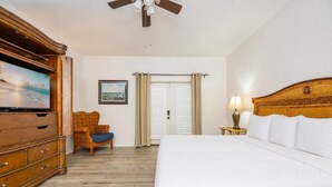 Quarto de hotel com cama, televisão, cómoda de madeira, cadeira, abajur e ventoinha no teto.