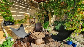 Uma área de estar ao ar livre com uma mesa de madeira e duas cadeiras, uma placa com a inscrição "Come in OPEN" e uma parede de pedra com plantas trepadeiras.