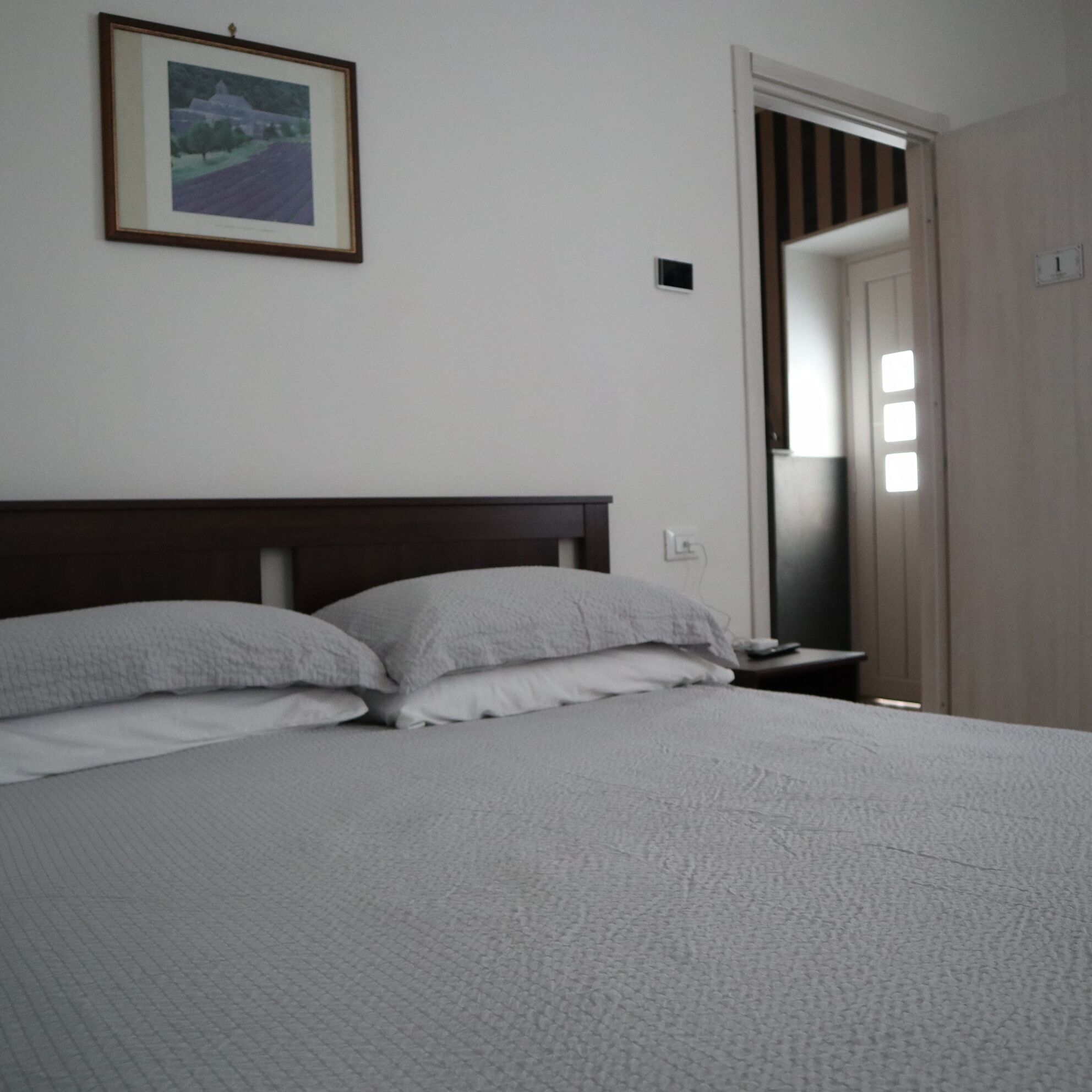 Quarto com cama, almofadas, mesa de cabeceira, quadro na parede e porta que dá para outro cômodo.