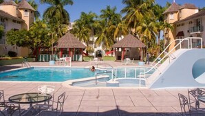 Área de piscina com espreguiçadeiras e um escorregador, cercada por palmeiras e edifícios com torres.