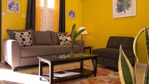 Uma sala de estar com sofá, poltrona e mesa de centro, com obras de arte nas paredes e uma janela com cortinas.