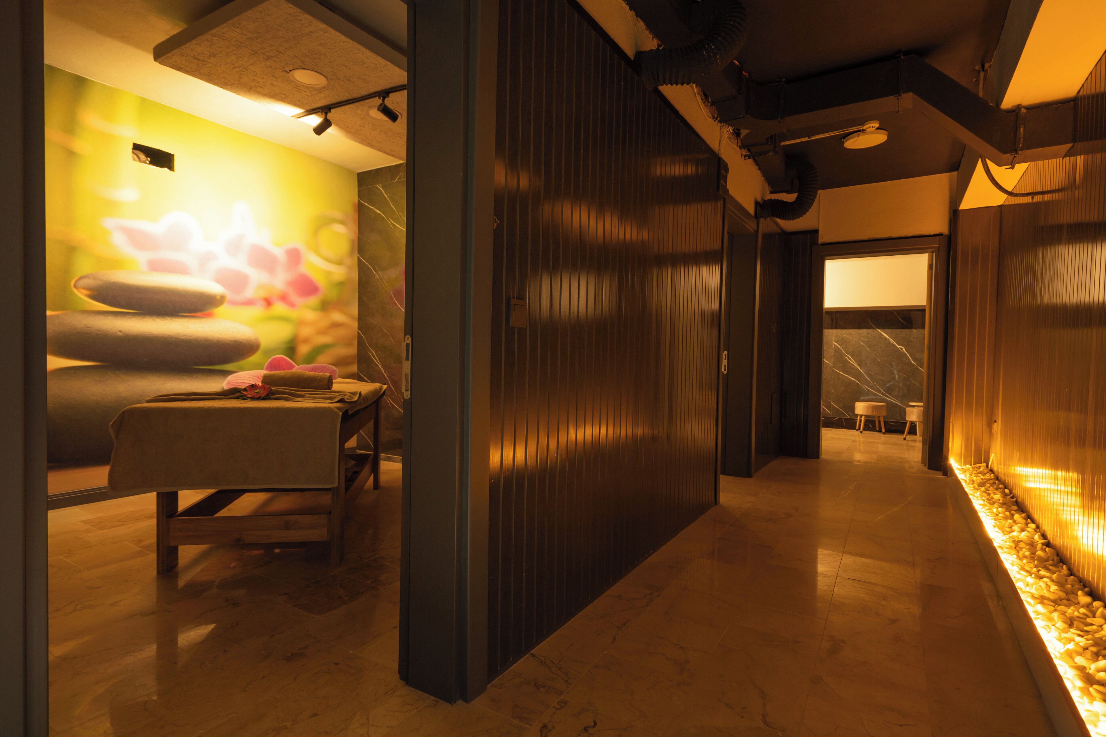 Uma área de spa com mesa de massagem, parede com painel floral e uma mesa de recepção ao fundo.