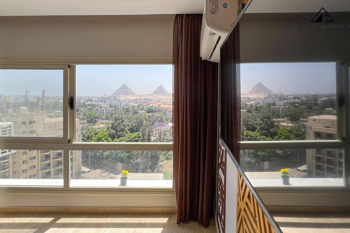 Quarto com grandes janelas que oferecem vista para as pirâmides e o horizonte urbano.