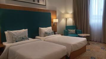 Quarto de hotel com duas camas, uma poltrona verde-água, uma mesinha redonda e uma luminária.