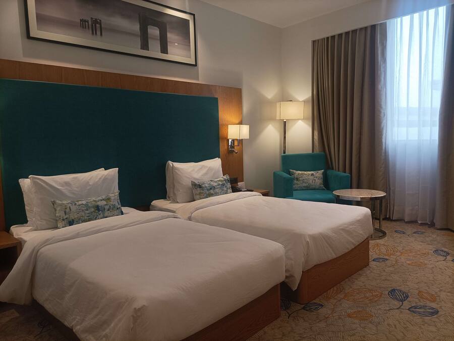 Quarto de hotel com duas camas, uma poltrona verde-água, uma mesinha redonda e uma luminária.