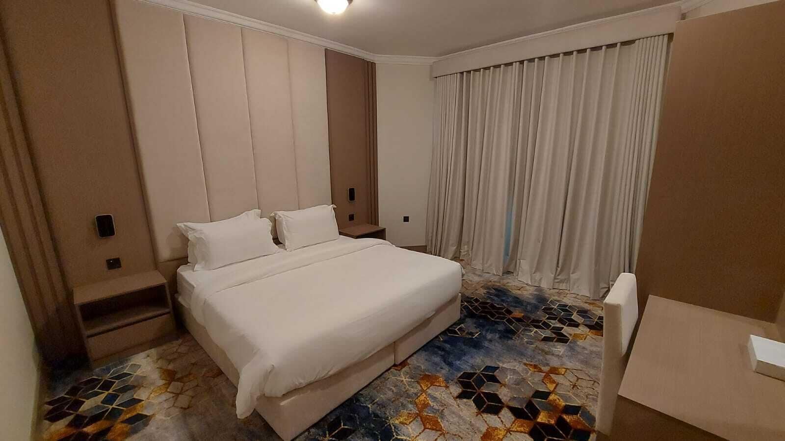 Quarto de hotel com cama grande, mesa de cabeceira, guarda-roupa e cortina.