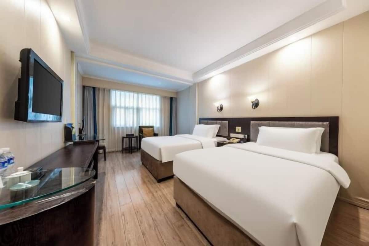 Quarto de hotel com duas camas, televisão, secretária com tampo de vidro e janela com cortinas.