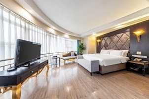 Quarto de hotel com cama, televisão sobre um console preto, escrivaninha com cadeira e mesa de cabeceira com telefone.