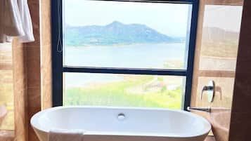 Um banheiro com banheira livre, uma janela com vista para um lago e montanhas, e um porta-toalhas.