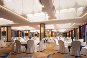 Salão de banquetes com mesas redondas e cadeiras, piso coberto por carpete e teto alto com luminárias.