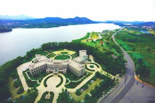 Vista aérea de um grande edifício histórico com layout simétrico, rodeado por jardins bem cuidados e um lago.
