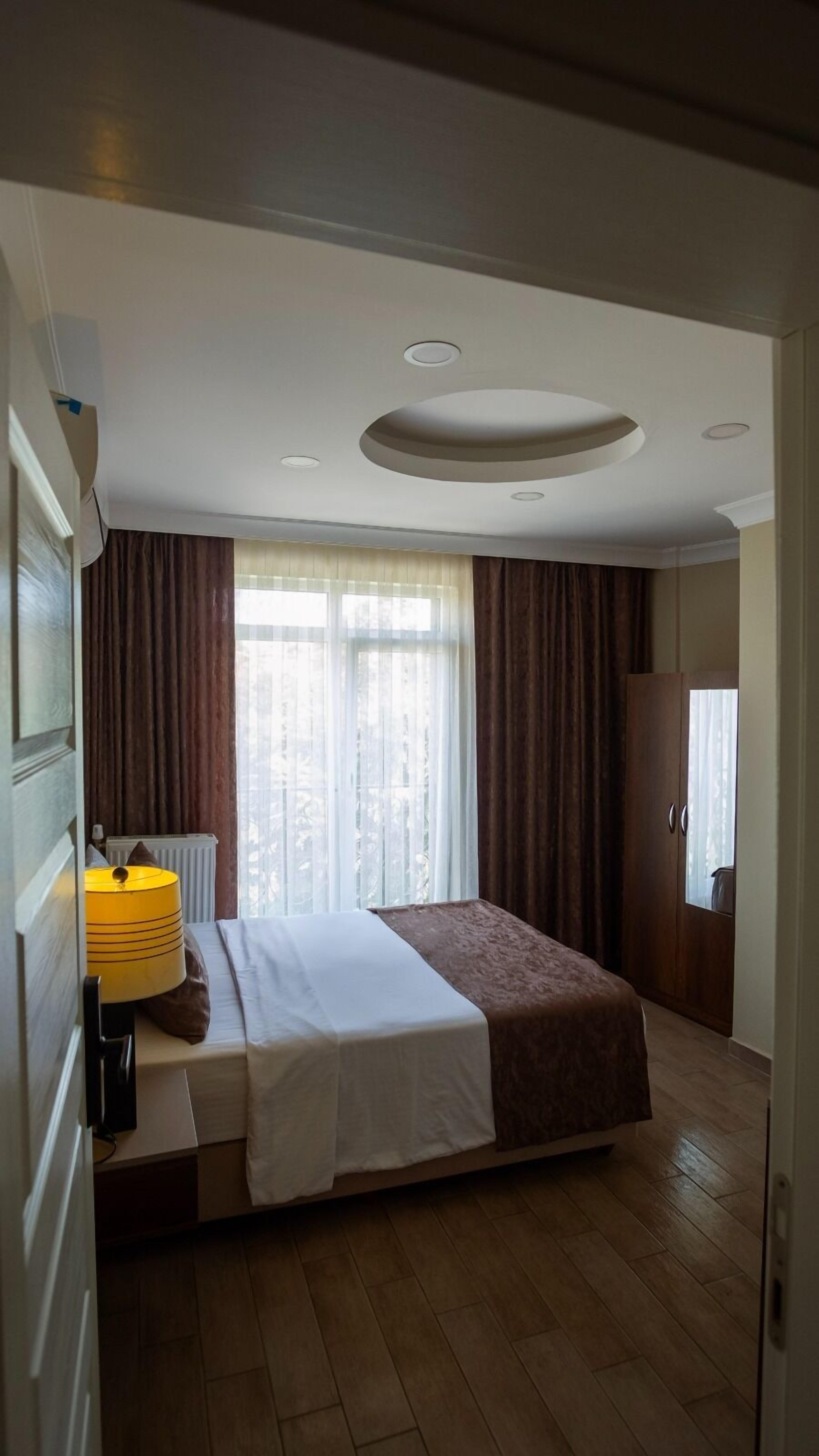 Quarto com cama, luminária de cabeceira, radiador e janela com cortinas.