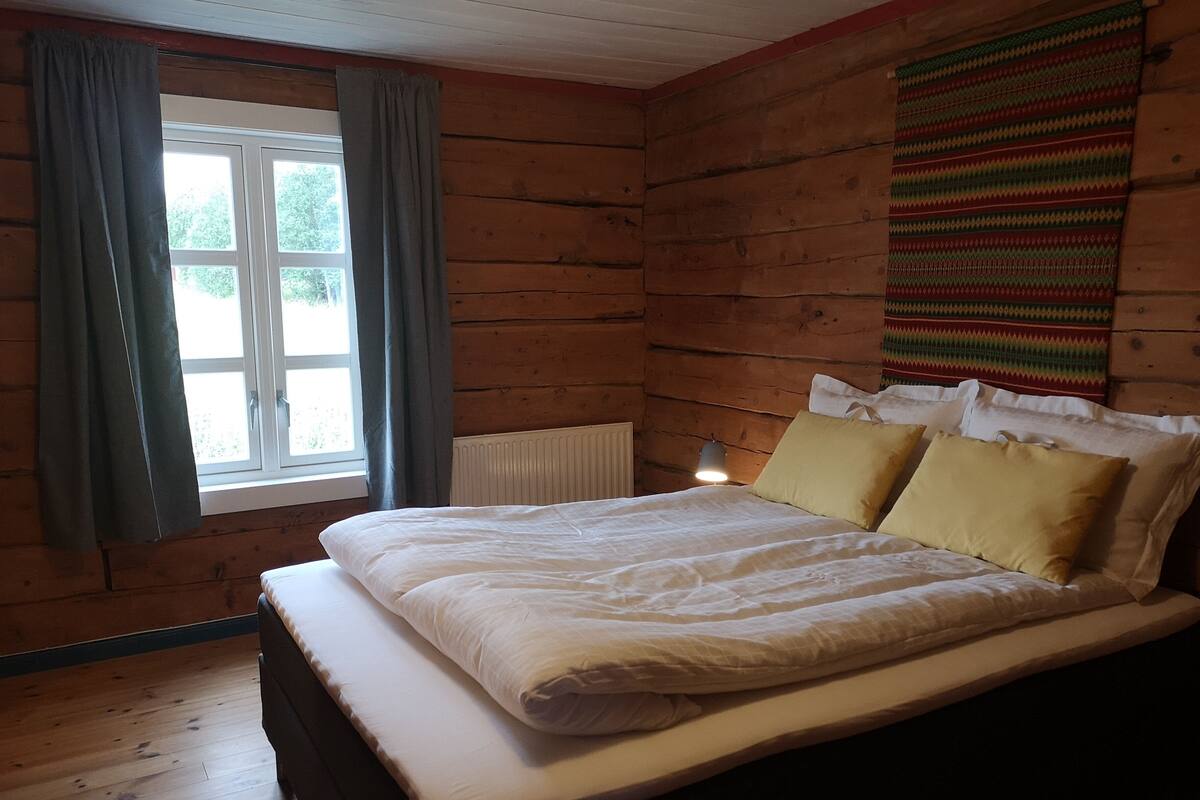 Quarto com painéis de madeira, cama, janela com cortinas, radiador e tapeçaria colorida.