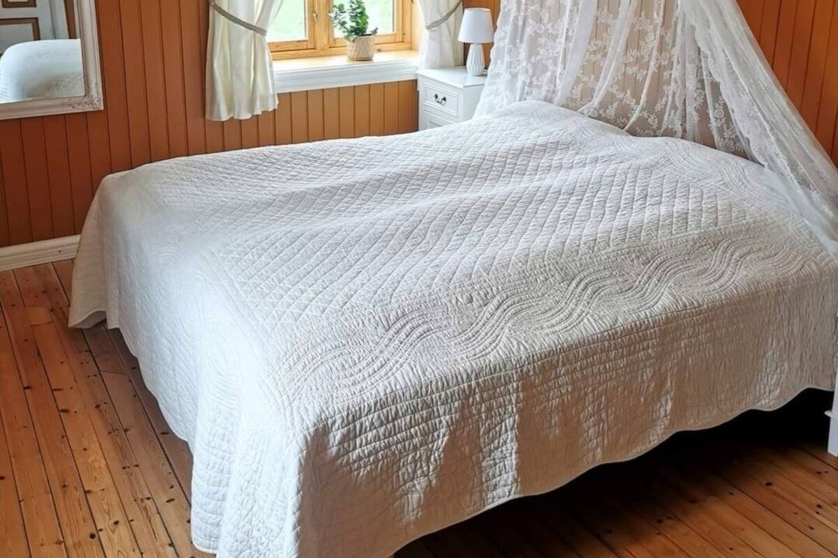 Quarto com cama, cortinas brancas, janela com vista, mesa de cabeceira com abajur e espelho na parede.
