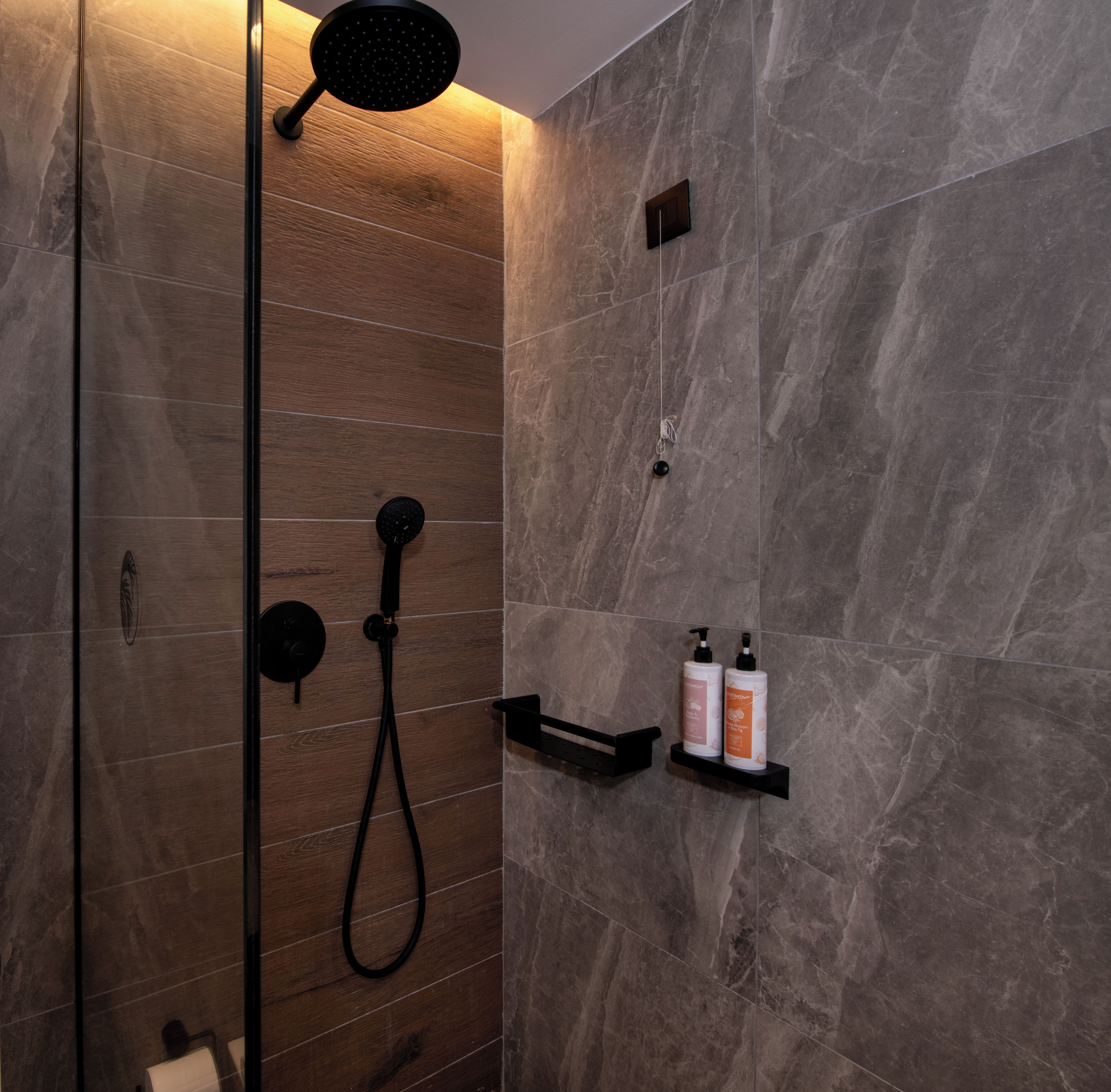 Um chuveiro moderno com um chuveiro de mão, painel de controle fixado na parede e uma prateleira com duas garrafas de shampoo.
