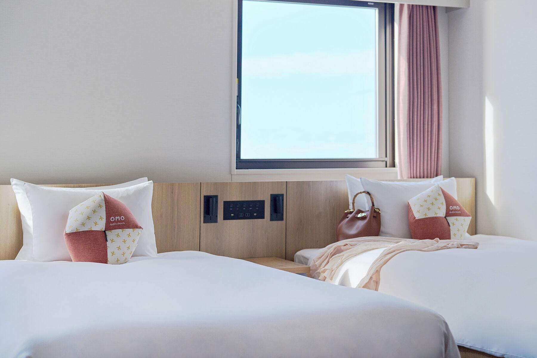 Quarto de hotel com duas camas, cada uma com travesseiro ostentando um logotipo e texto, uma janela com vista e um painel de controle na parede.