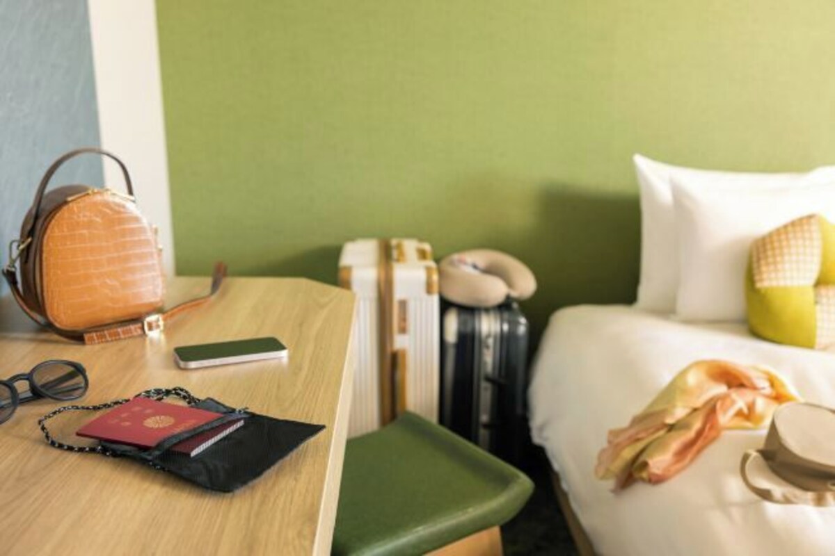 Quarto de hotel bem arrumado com cama, almofadas, mesa de cabeceira com uma bolsa, óculos e uma pequena sacola.