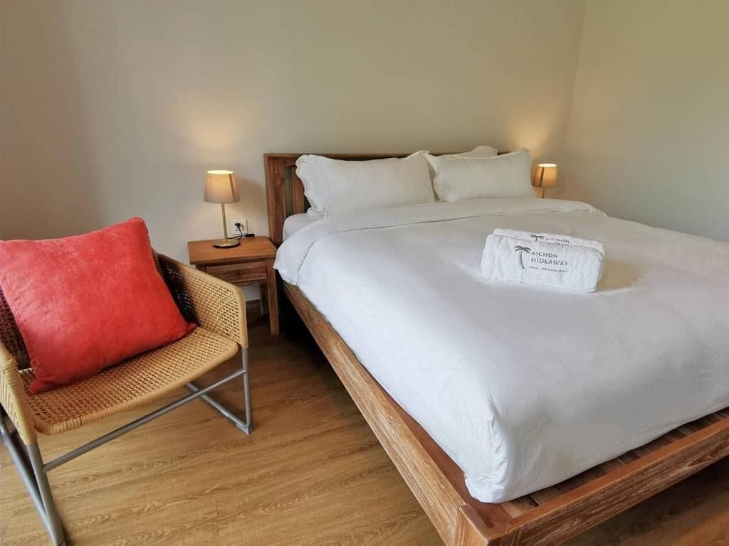 Cama bem arrumada com lençóis brancos e toalha, mesa de cabeceira de madeira com abajur e cadeira de vime com almofada vermelha.