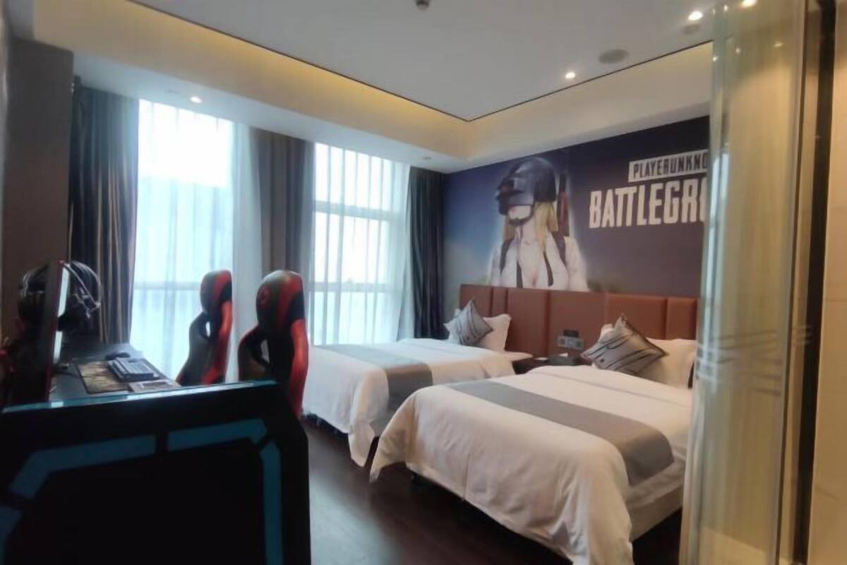 Quarto de hotel com duas camas, uma escrivaninha com cadeira, uma cadeira gamer e um pôster grande na parede.