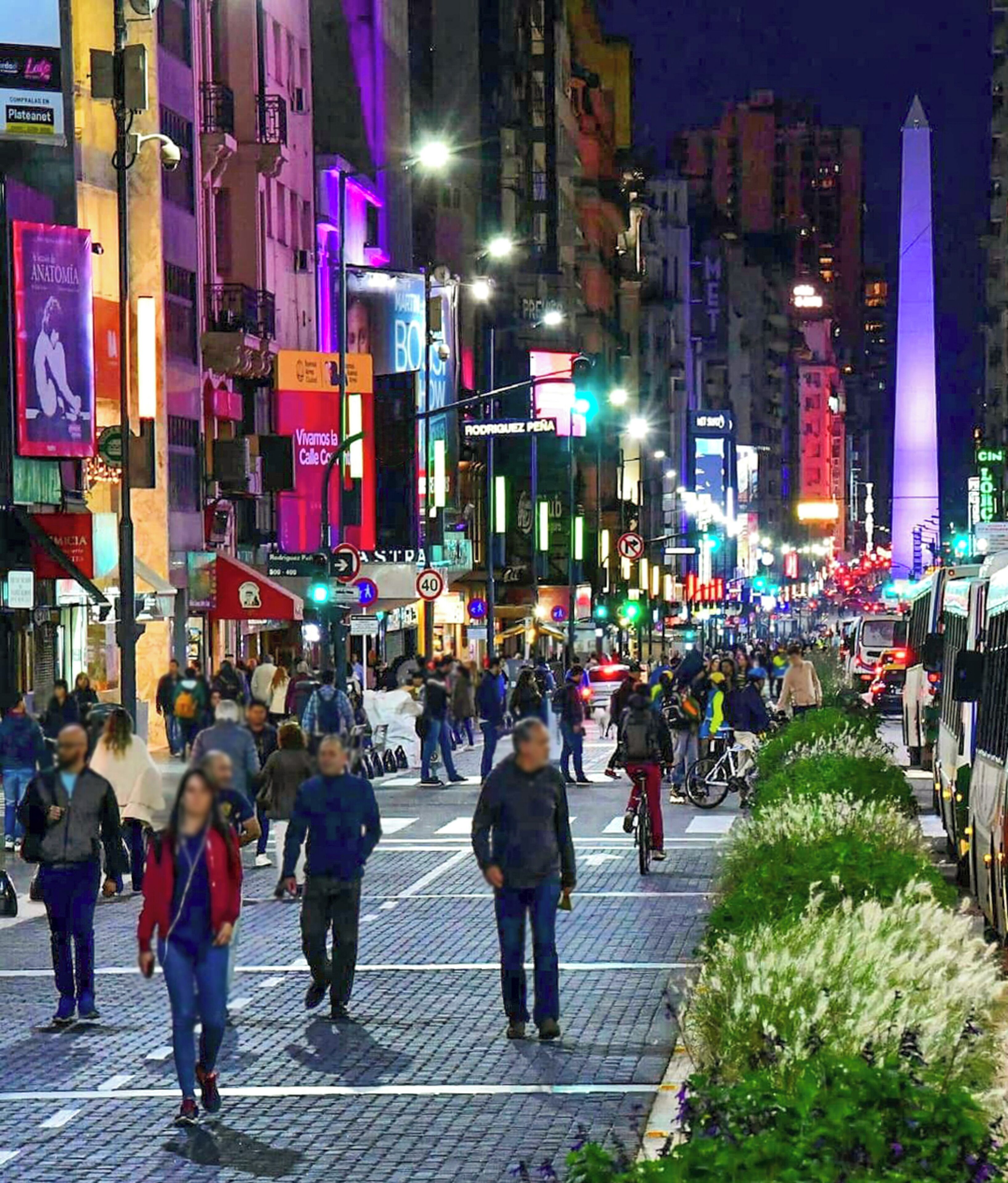 Rua urbana movimentada à noite, com pedestres e veículos, iluminada por letreiros de néon coloridos e postes de luz.